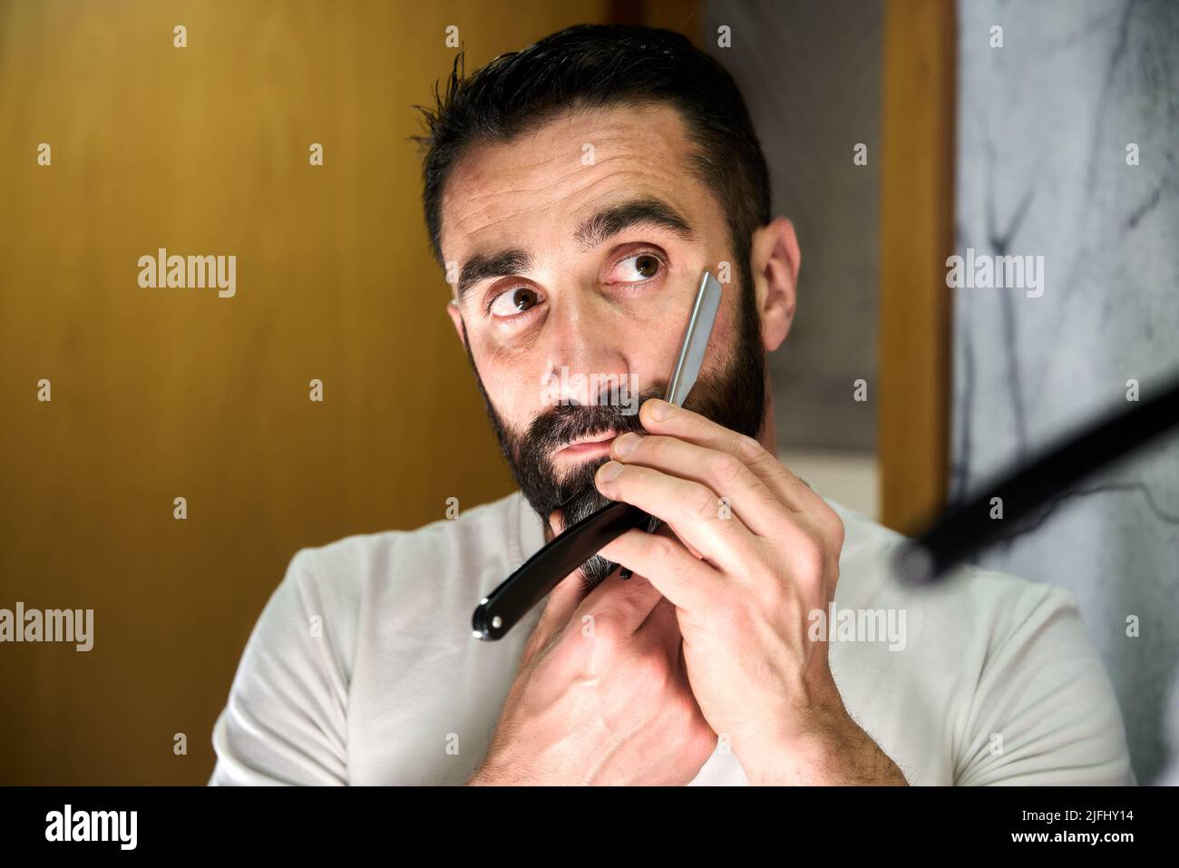 Young man with a beard shaving with a traditional knife in front of the ...