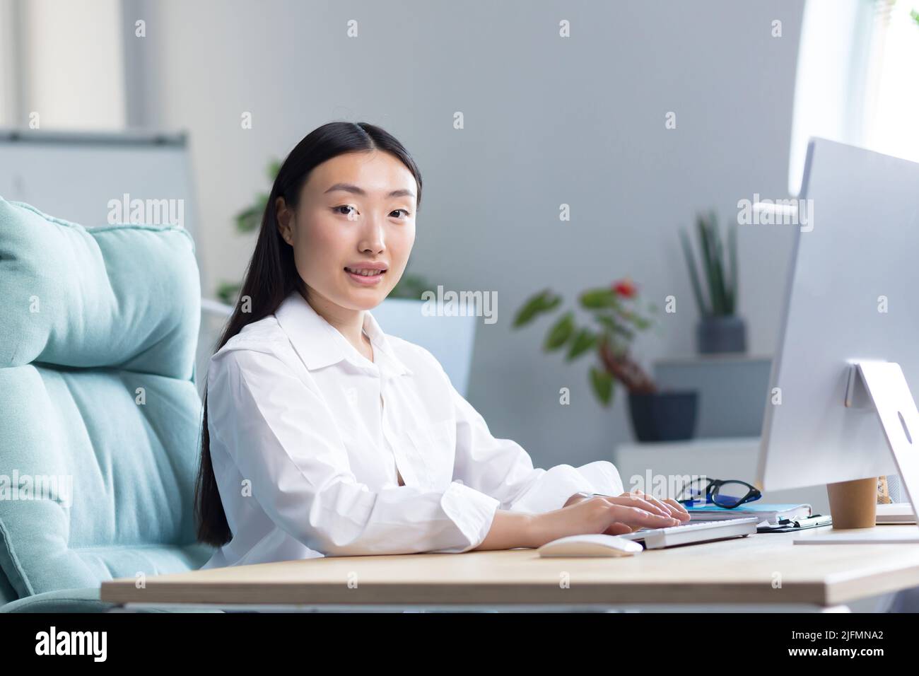Beautiful Asian woman works at the computer in the office, looks at the ...