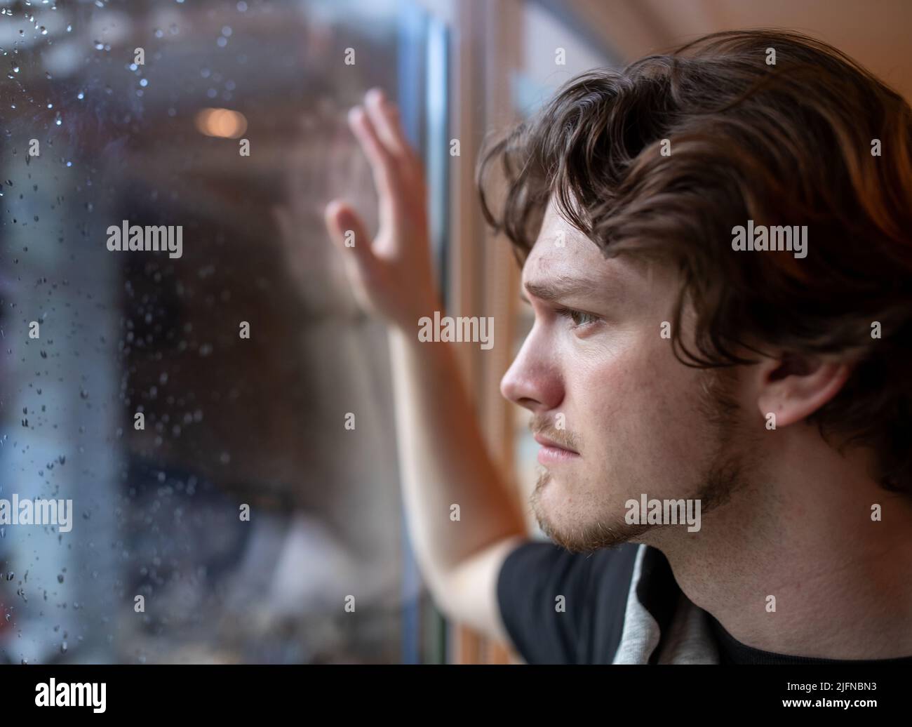 Worried young man looking through window at home on rainy day Stock ...