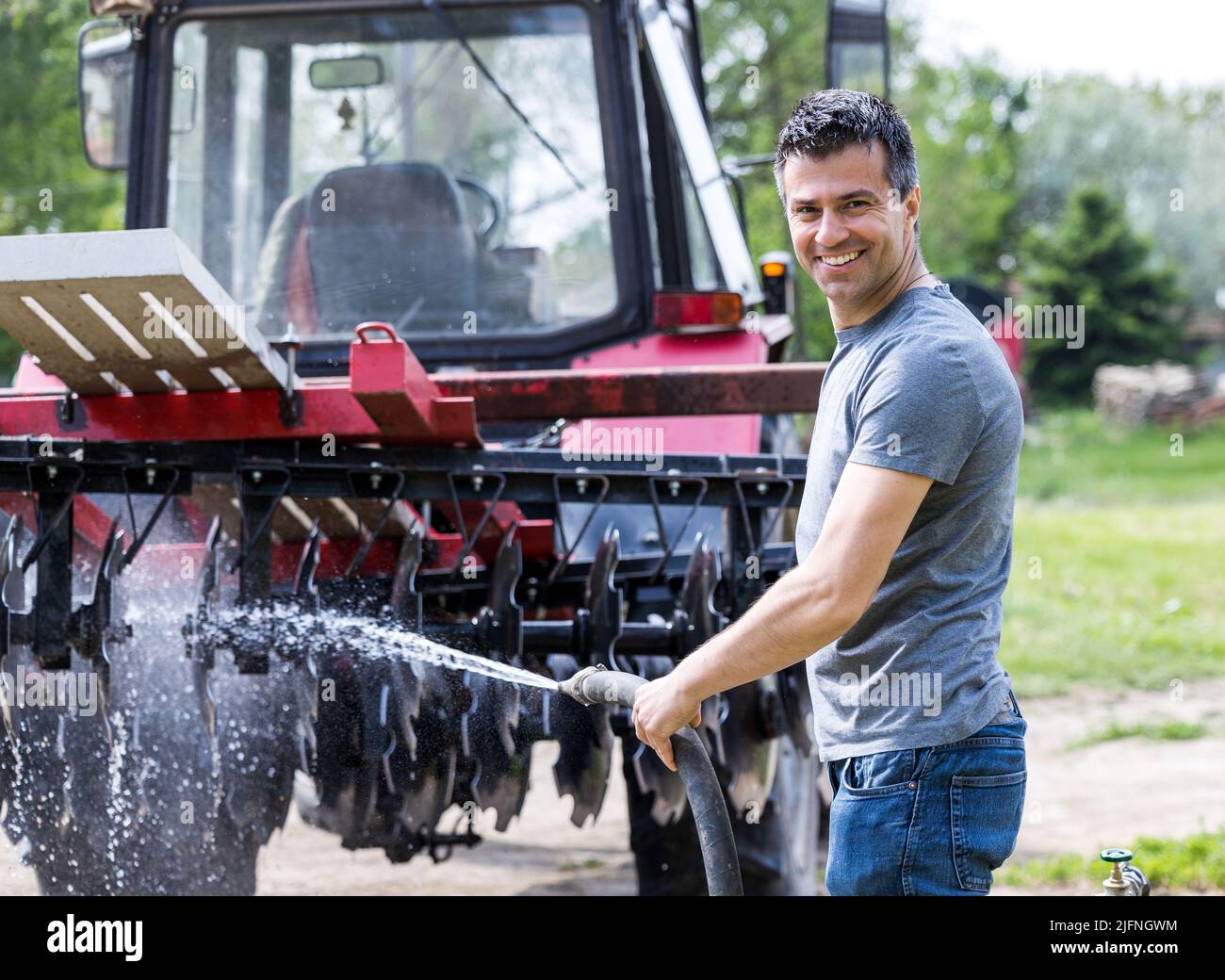 Handsome satisfied man cleaning tractor and equipment with water stream ...