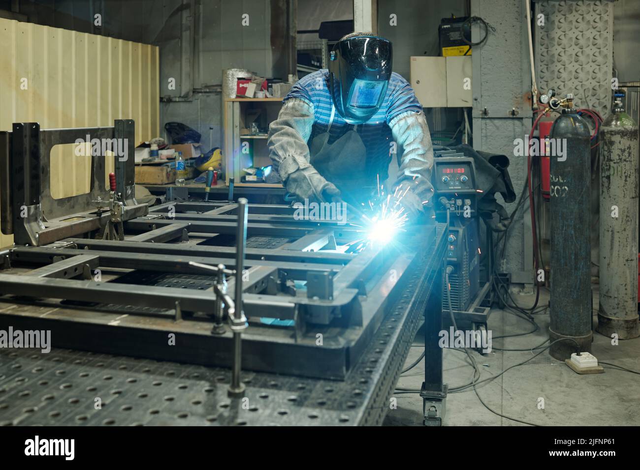 Young male specialist in protective workwear welding metallic parts of ...