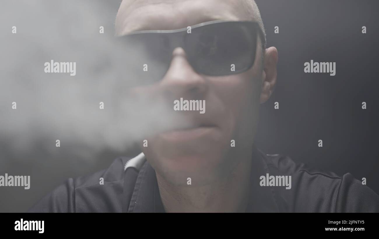 Man in black glasses exhales smoke. Action. Bandit blows smoke in dark ...