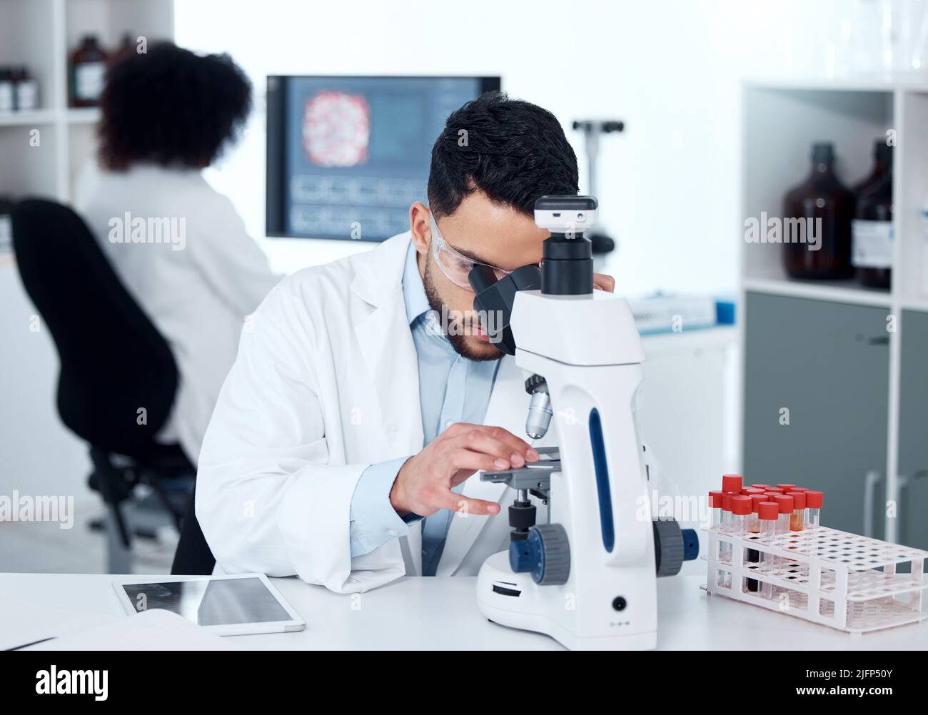 A serious medical scientist sitting at a desk and using a microscope to ...