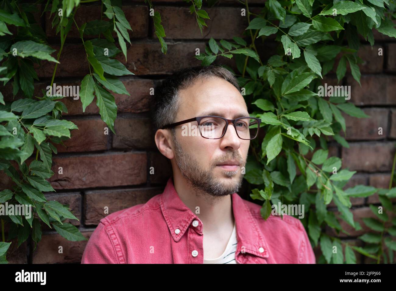 Headshot portrait of smiling 30s caucasian millennial man in glasses sit at street on brick wall ...