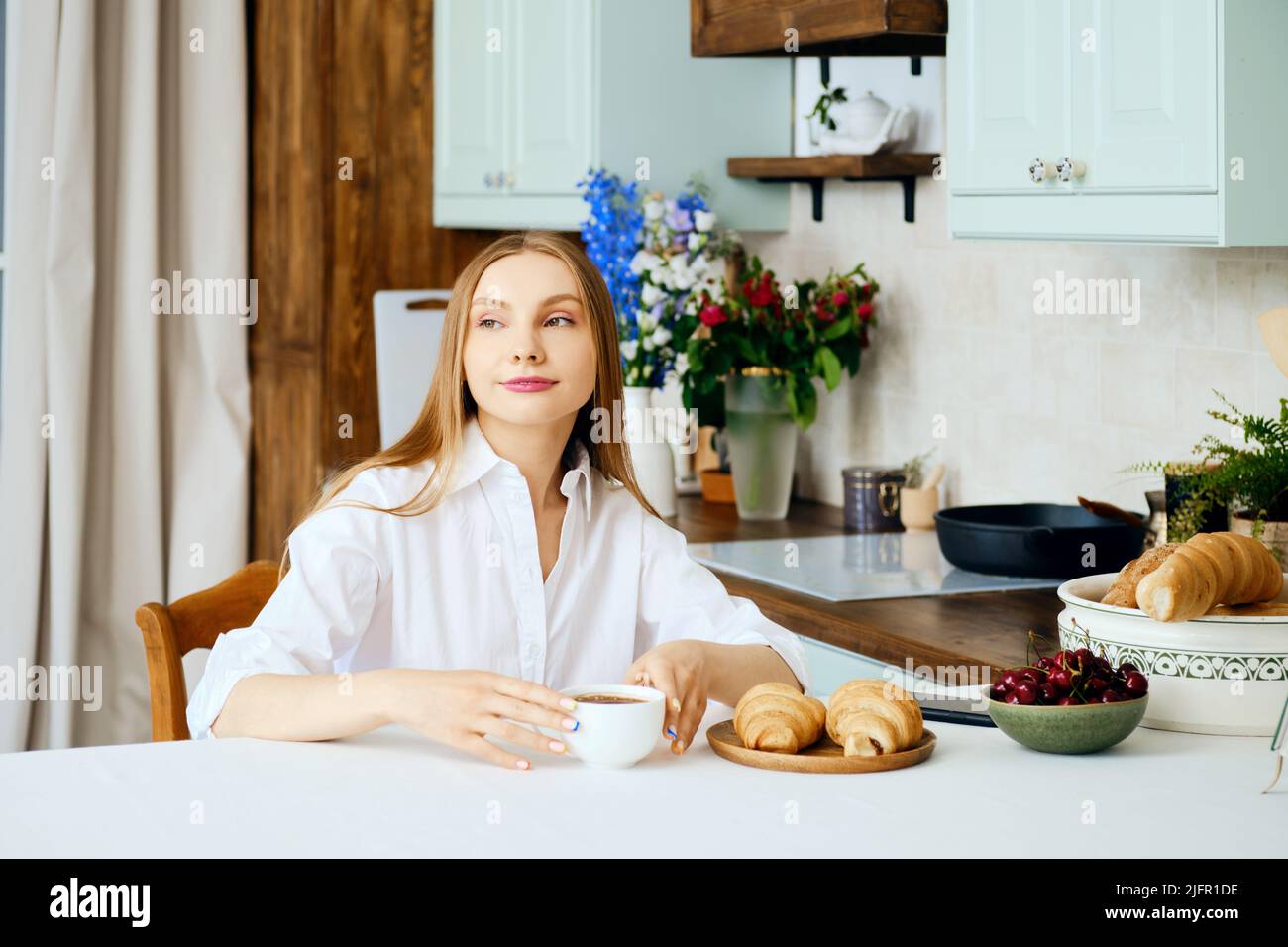 Positive young woman sits behind the kitchen table with cup of coffee ...