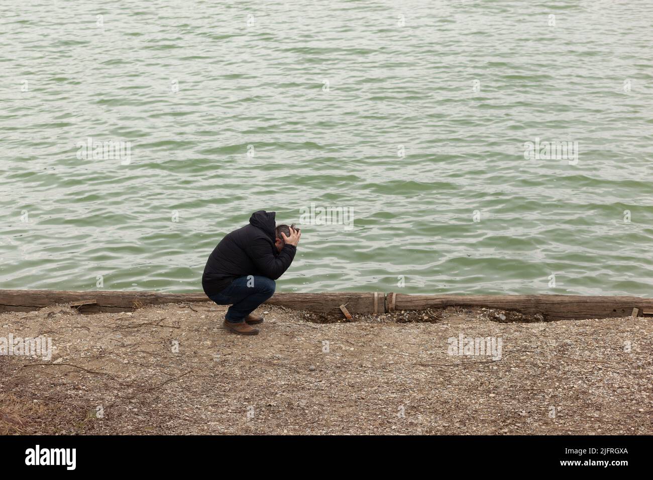 Man crouching by the water, hands on heads, desperate and afraid Stock ...