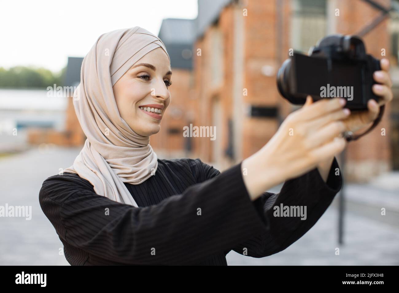Young female muslim blogger in trendy clothes and hijab walks on city street, records herself on ...