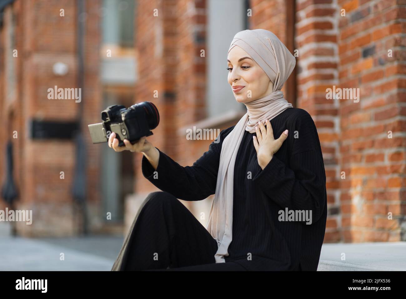 Young female muslim blogger in trendy clothes and hijab sitting on stairson city street, records ...