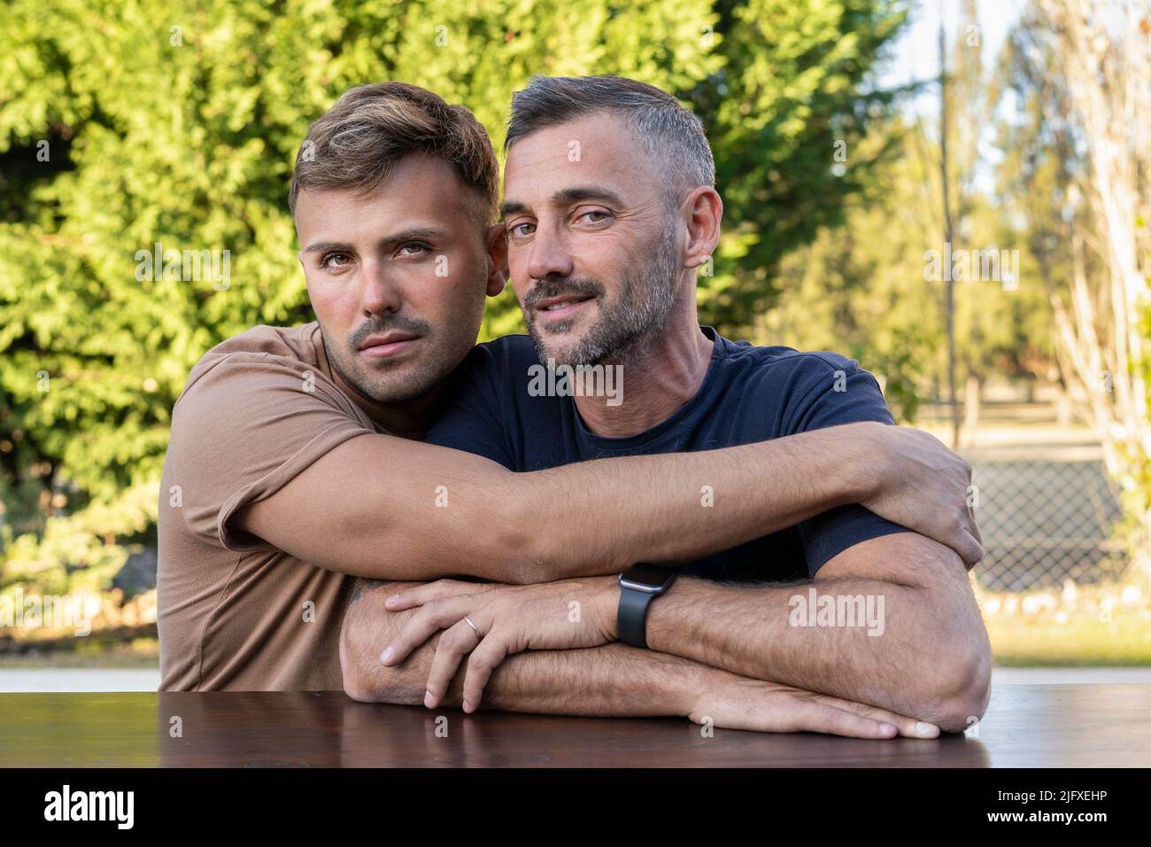 A young man hugging his husband outdoors. Both are looking at camera ...