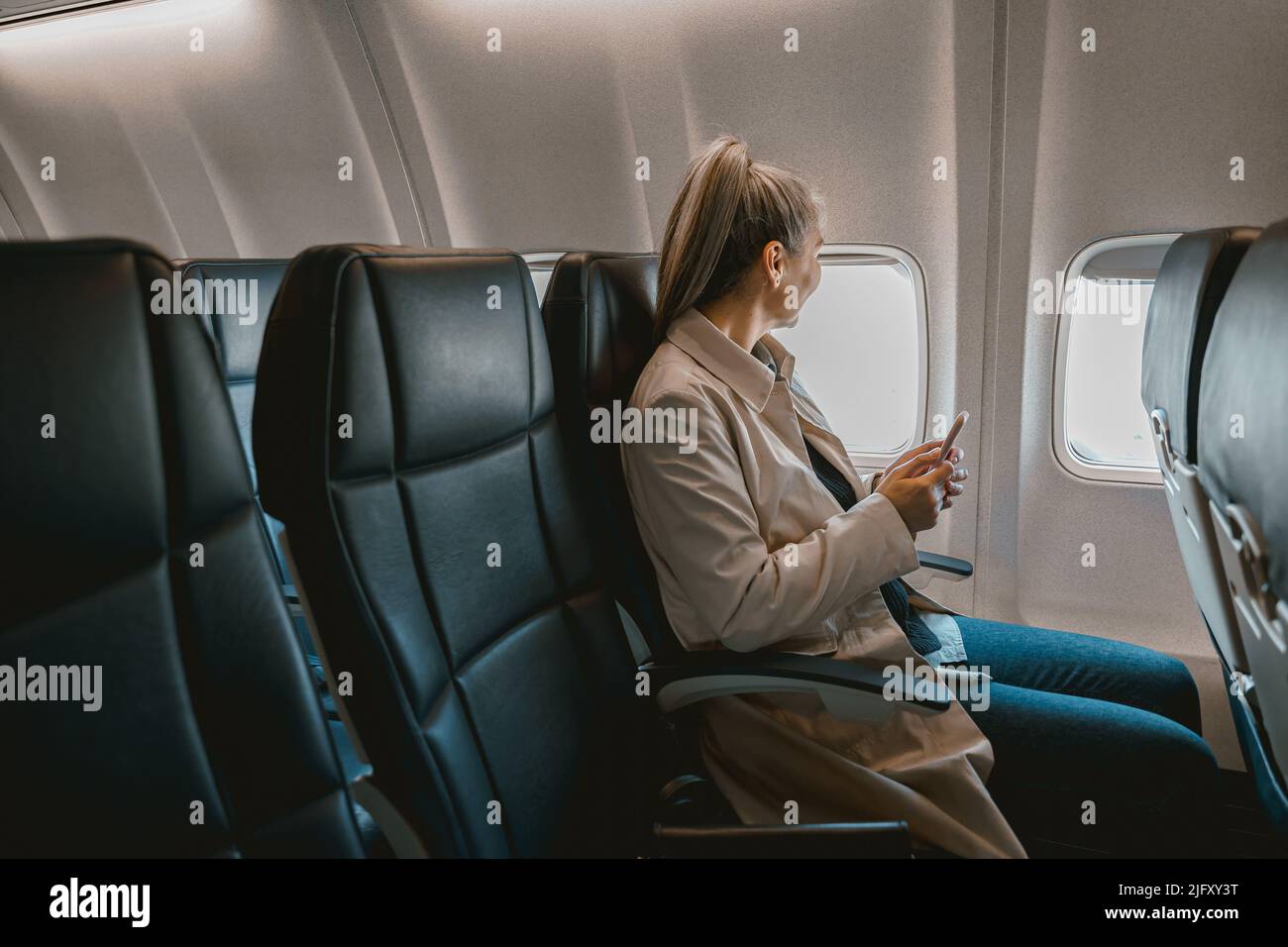 Beautiful Asian woman passenger sitting in the airplane and holding phone and looking window ...