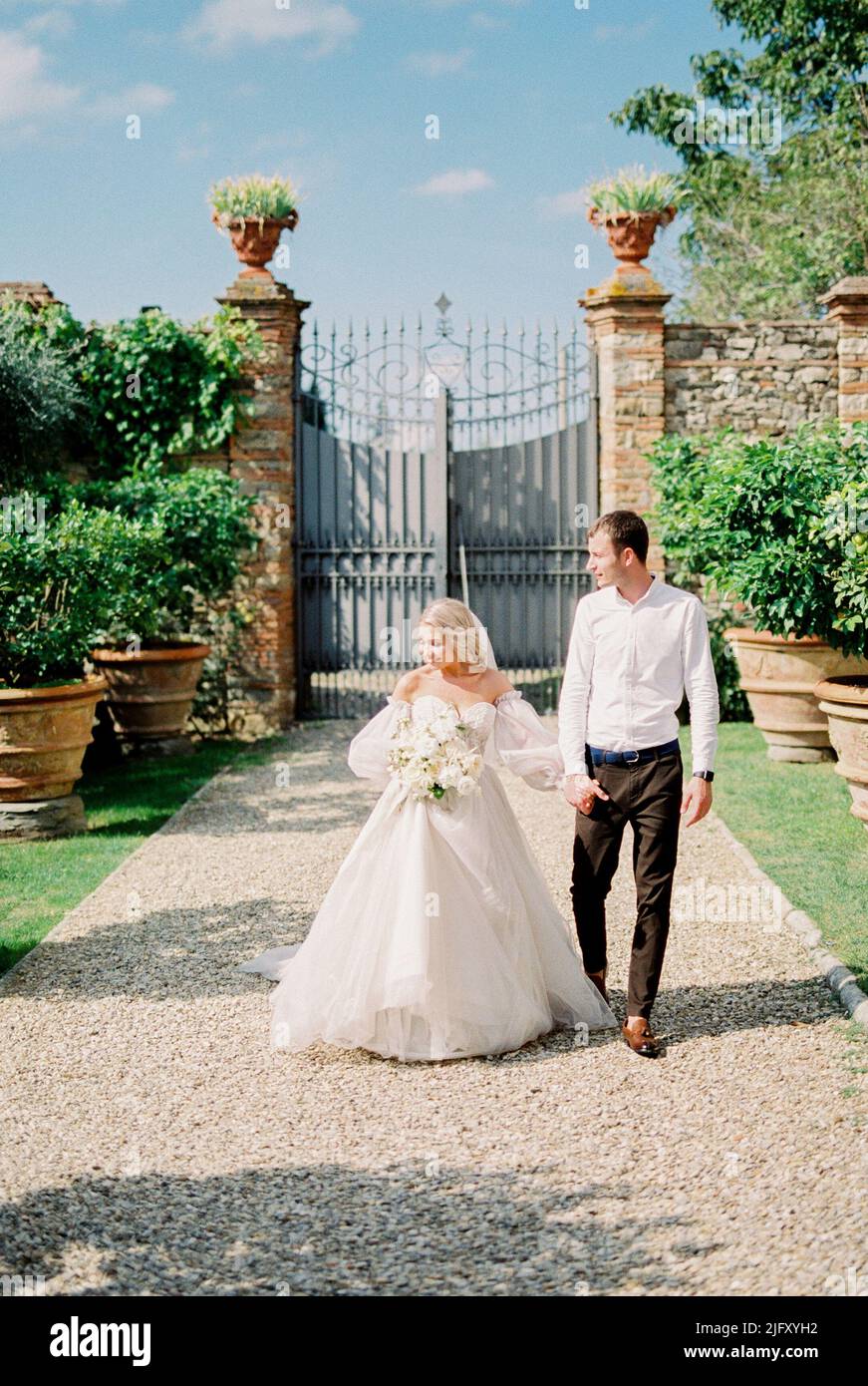 Bride and groom walk along the gravel path in the park holding hands ...