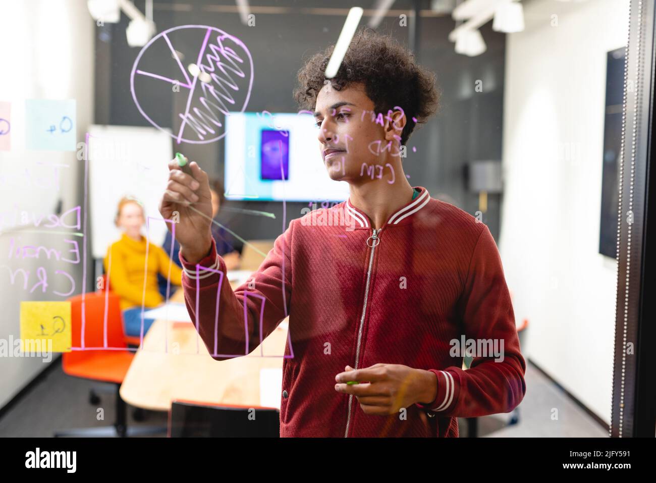 Young biracial male app developer drawing chart on glass in board room ...