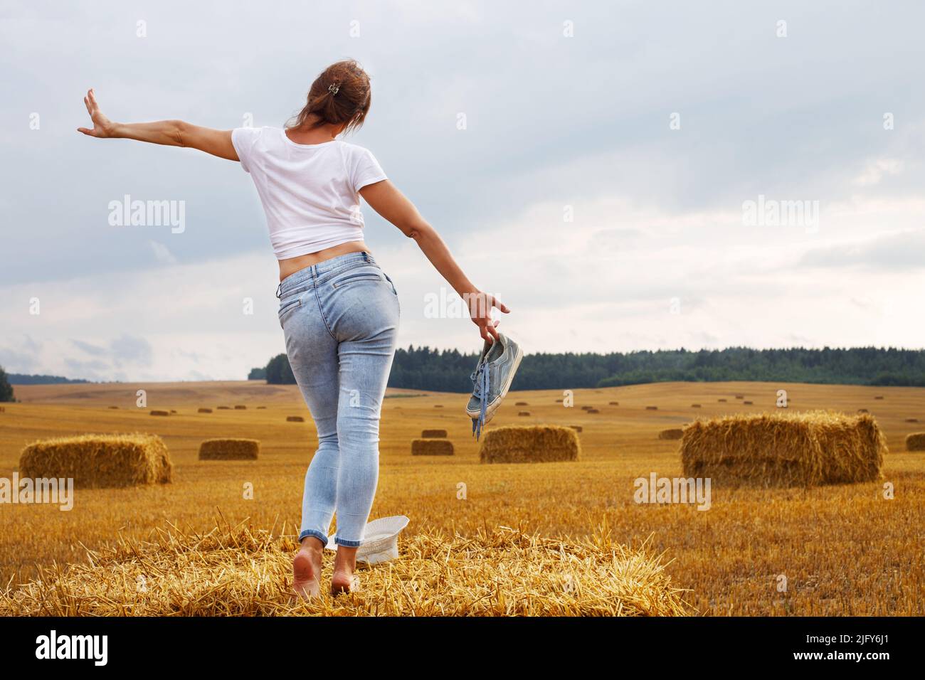 barefoot girlwith sneakers in hand stands on a haystack on a bale in the agricultural field ...