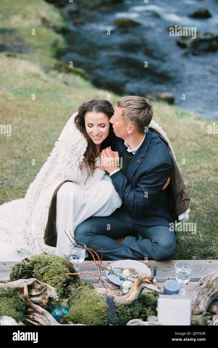 Groom kisses bride in a blanket at the laid table on the river bank ...