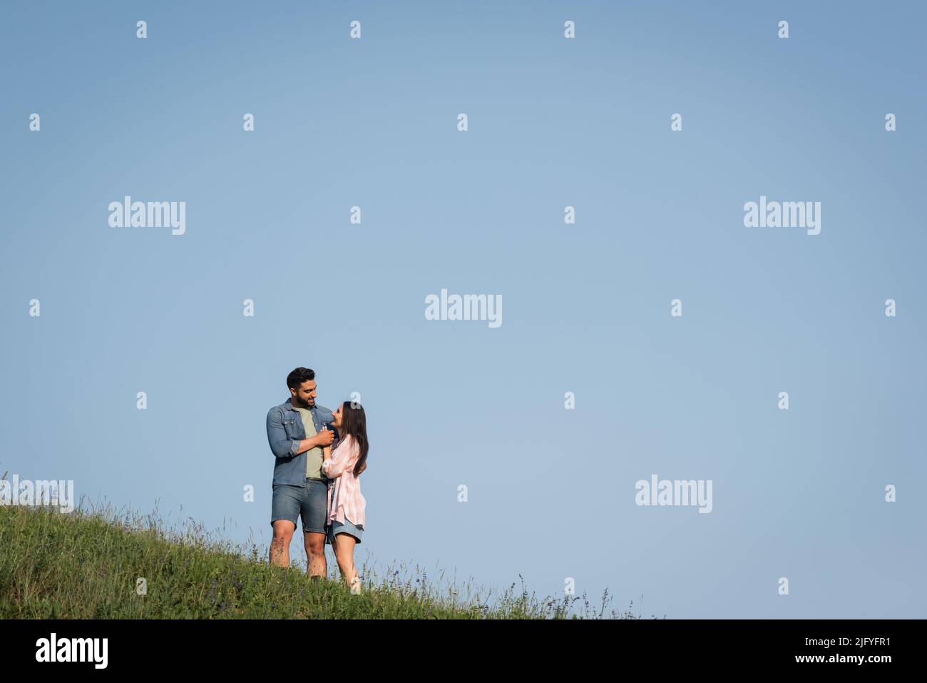 romantic couple looking at each other and embracing in field under blue ...