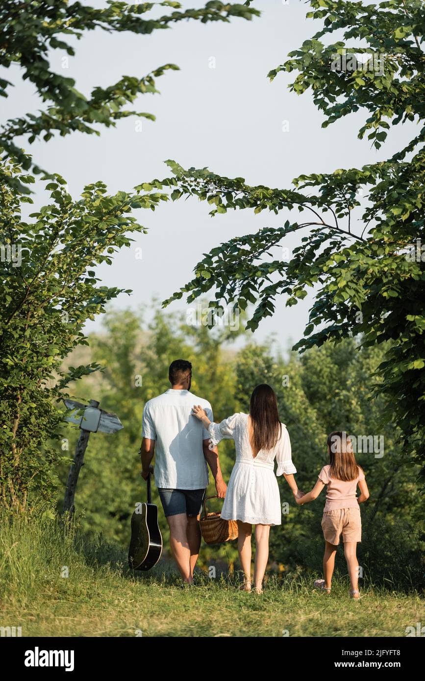 back view of family with wicker basket and acoustic guitar walking in ...