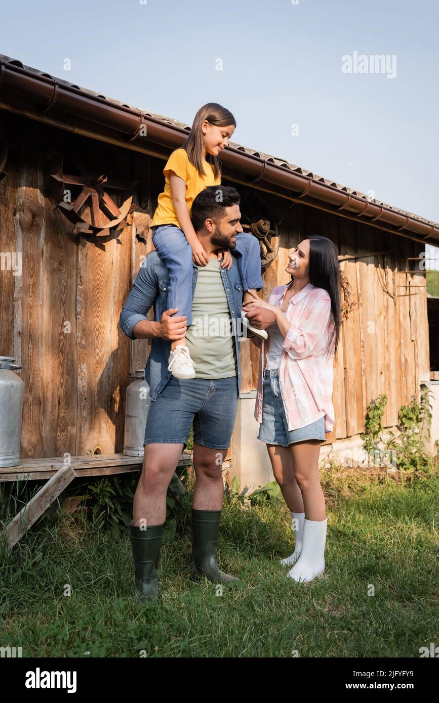 cheerful woman looking at husband piggybacking daughter near wooden ...