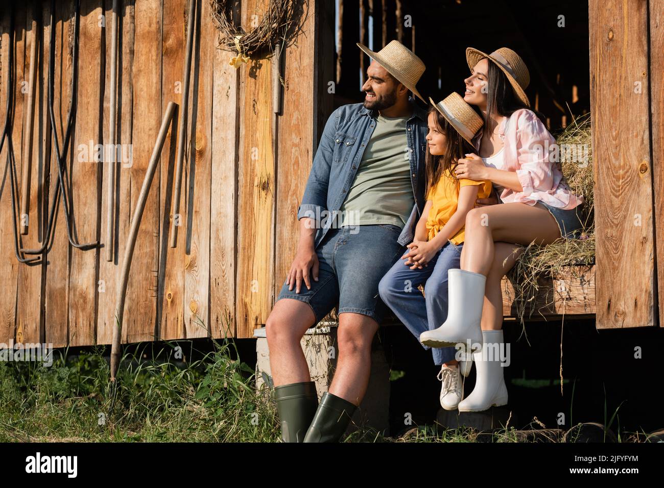 family of farmers in straw hats sitting on hay in wooden barn and ...