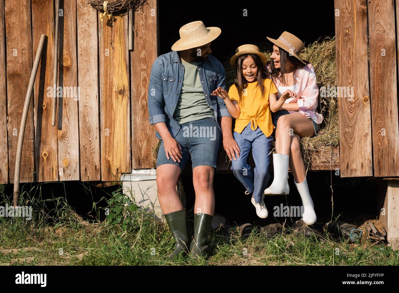 happy child in straw hat talking near parents on hay in wooden barn ...
