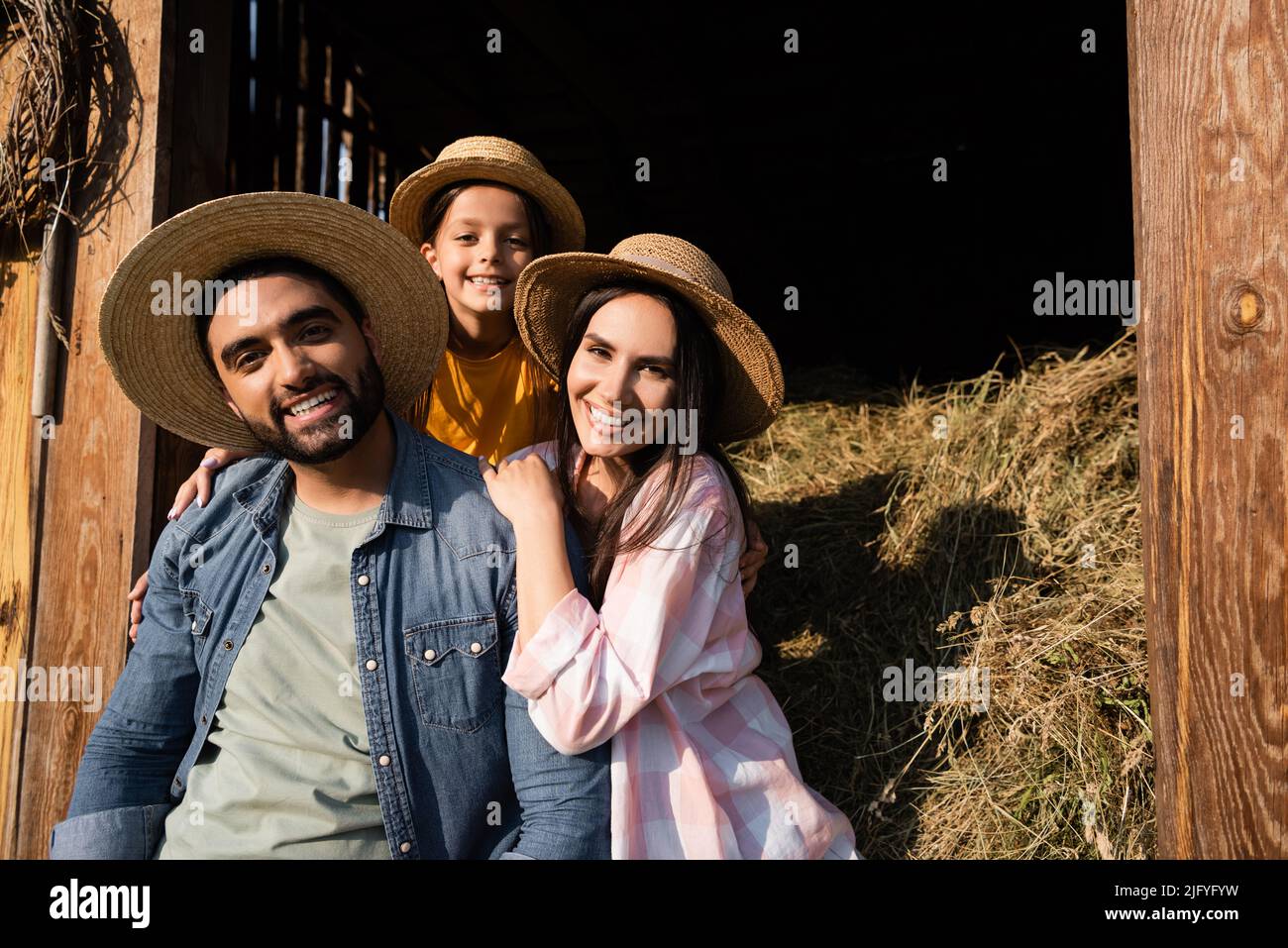 happy farm family in straw hats looking at camera near barn with hay ...