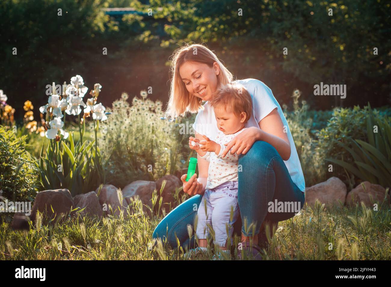 Smiling young mother hugs her baby daughter, who is playing with soap bubbles. Summer park on ...