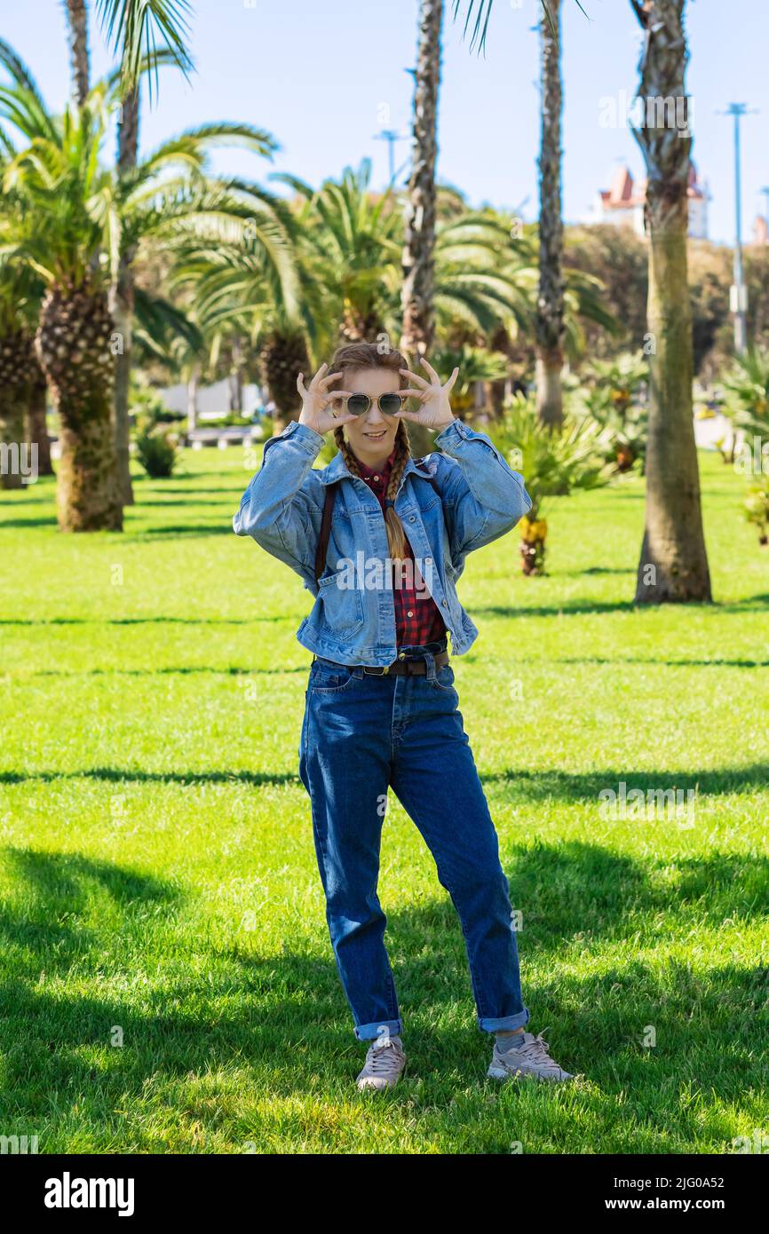 A woman standing on green lawn near palm trees holds on to sunglasses ...