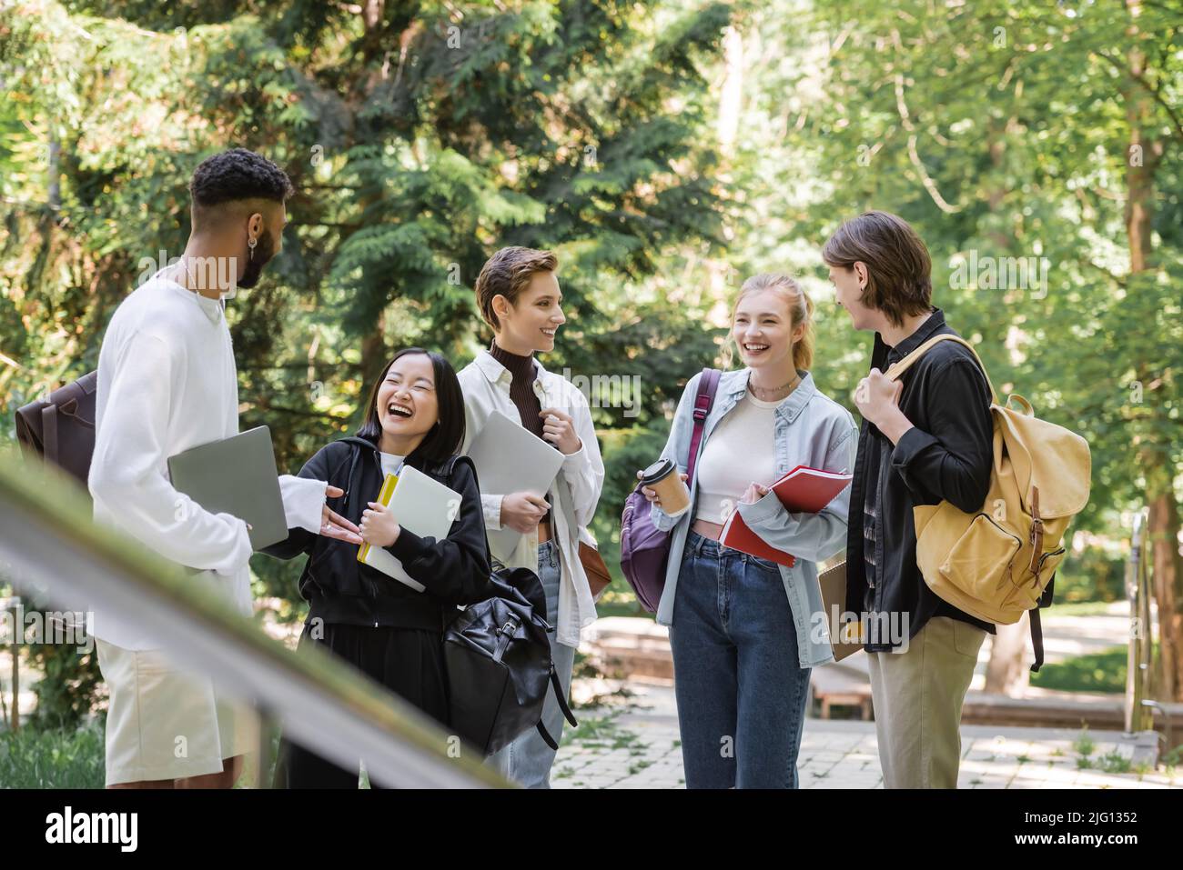 Cheerful interracial students with laptops and coffee to go in summer ...
