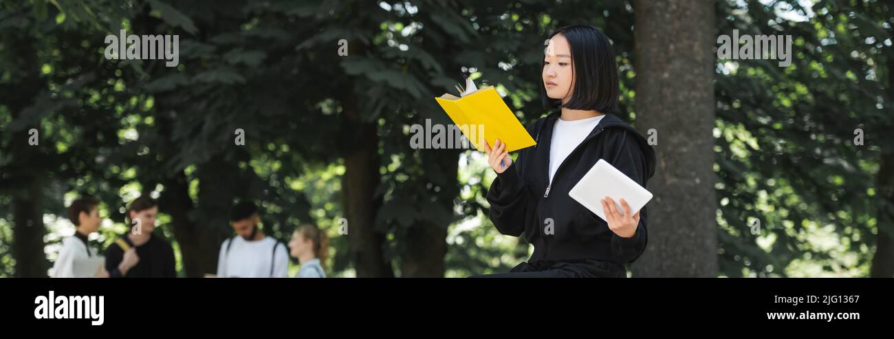 Asian student holding digital tablet and reading book in park, banner ...
