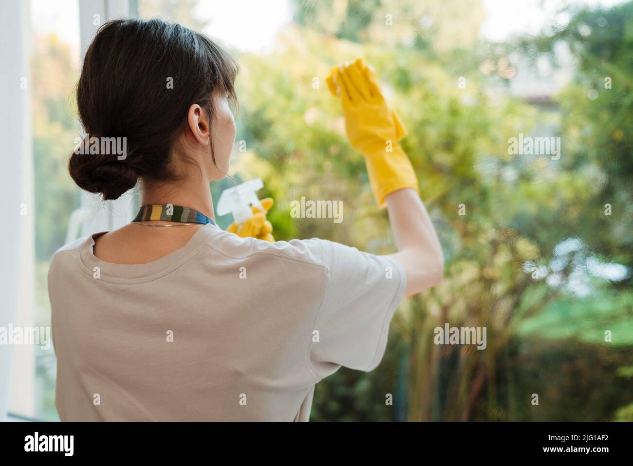White young woman in gloves washing window with cleaning spay at home ...