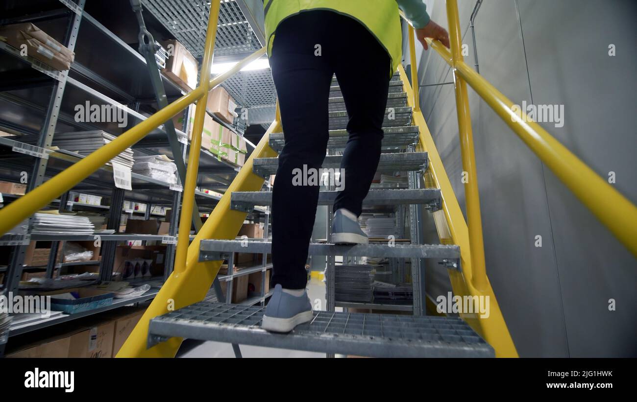 Rear view of a woman in uniform climbing up stairs inside warehouse ...