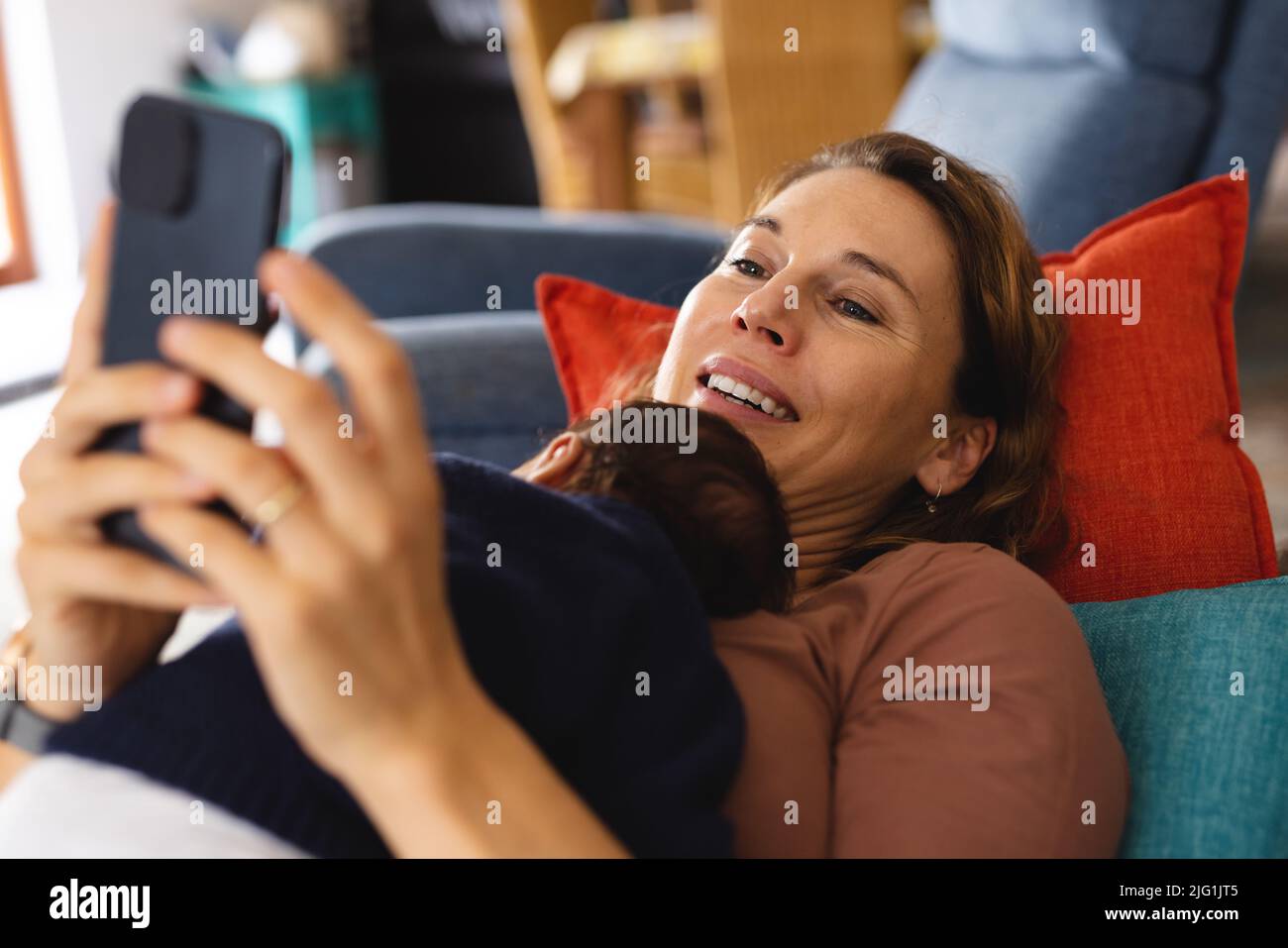 Smiling mid adult caucasian mother using smart phone while lying with ...