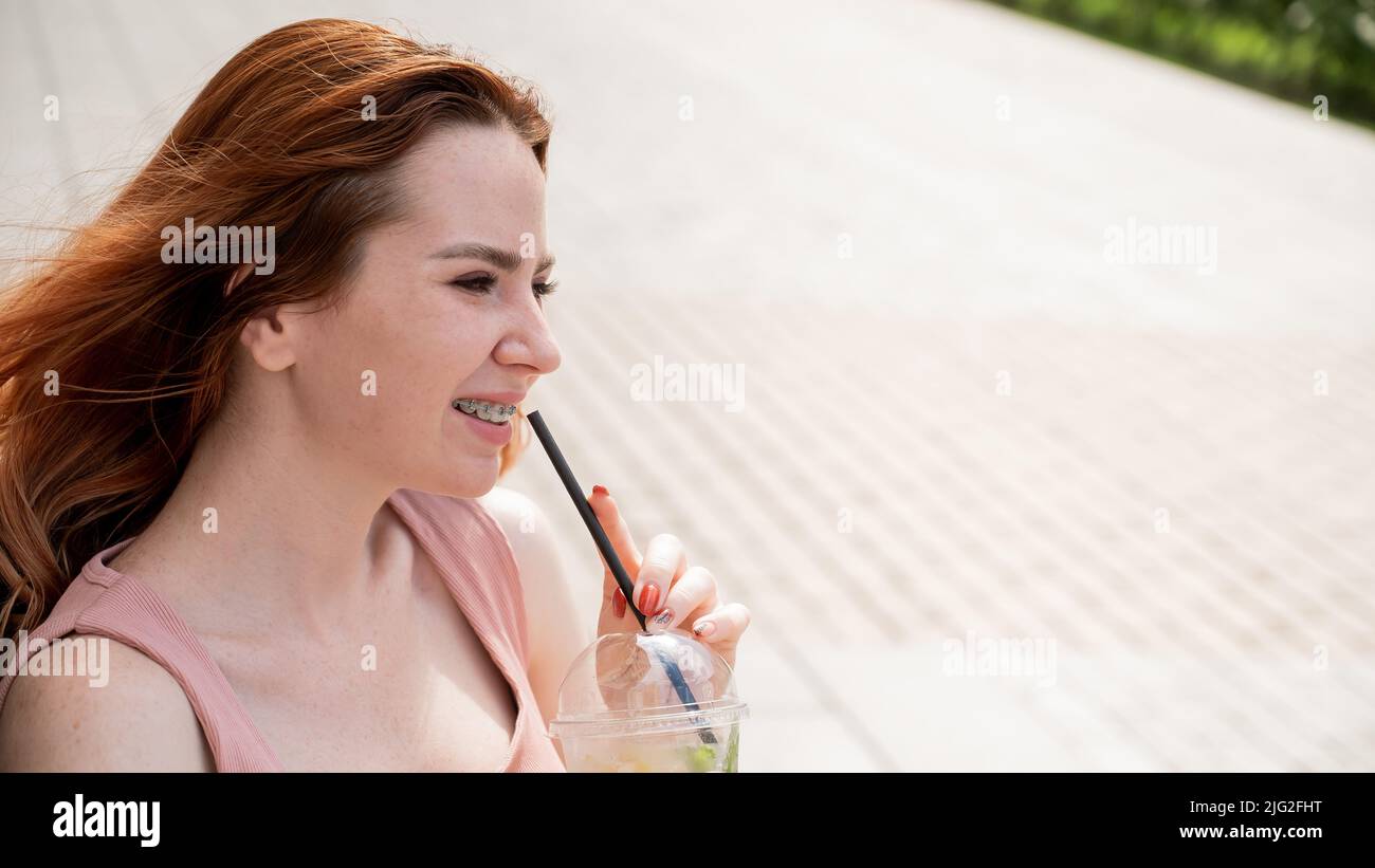 Young beautiful red-haired woman with braces drinks cooling lemonade ...