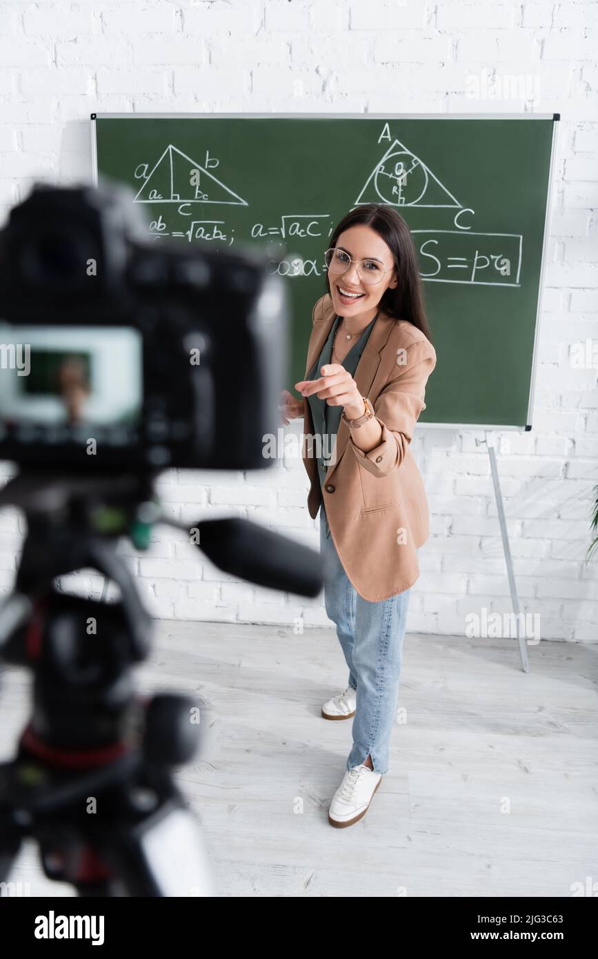 Positive teacher pointing at blurred digital camera and chalkboard in ...