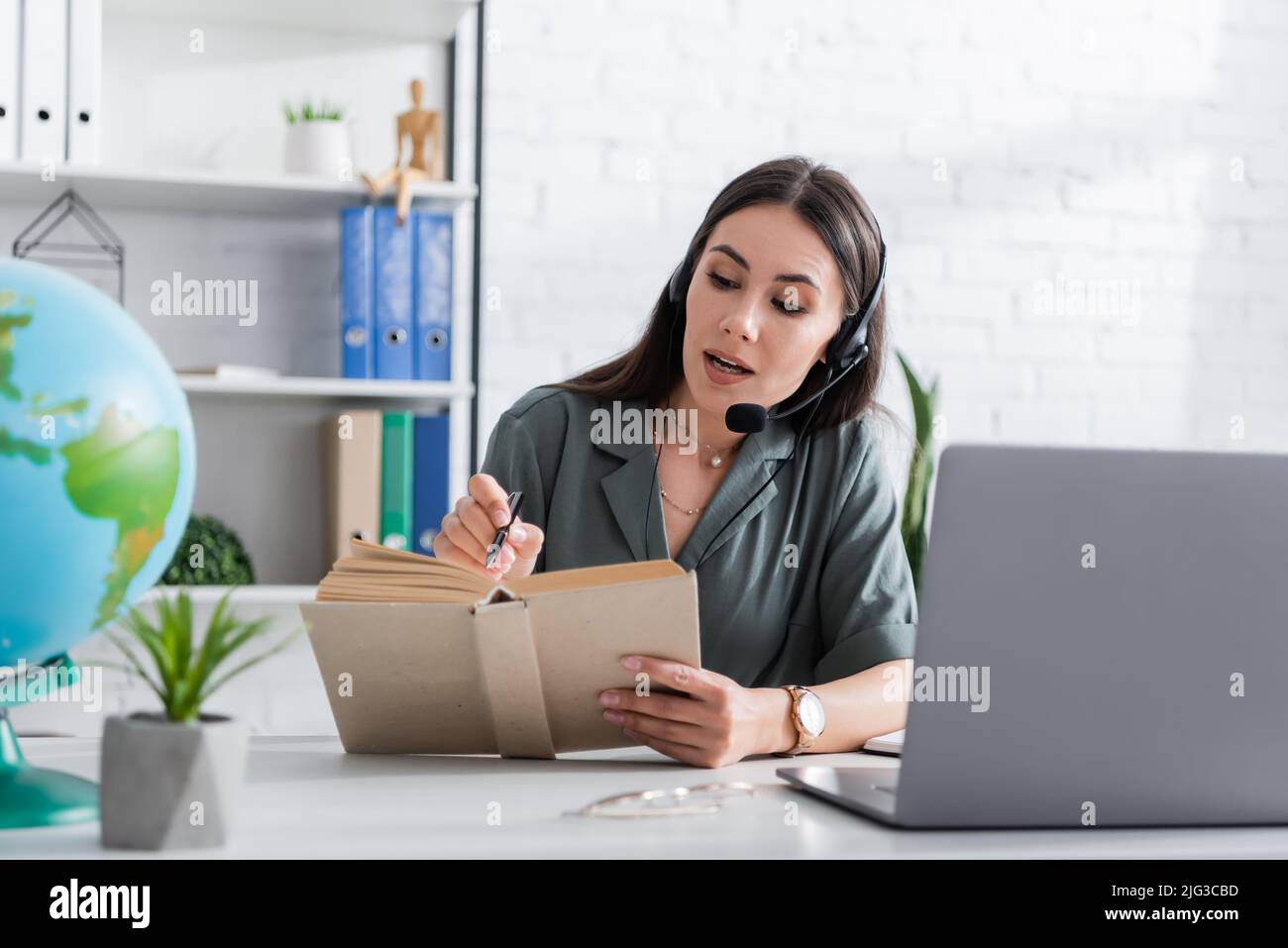 Teacher in headset holding book during online lesson on laptop in ...