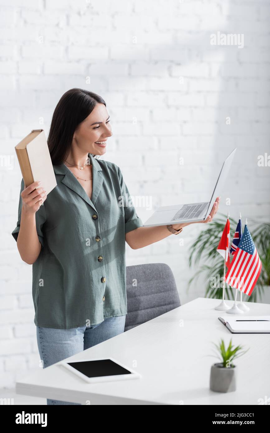 Cheerful teacher holding book and laptop near flags and digital tablet ...