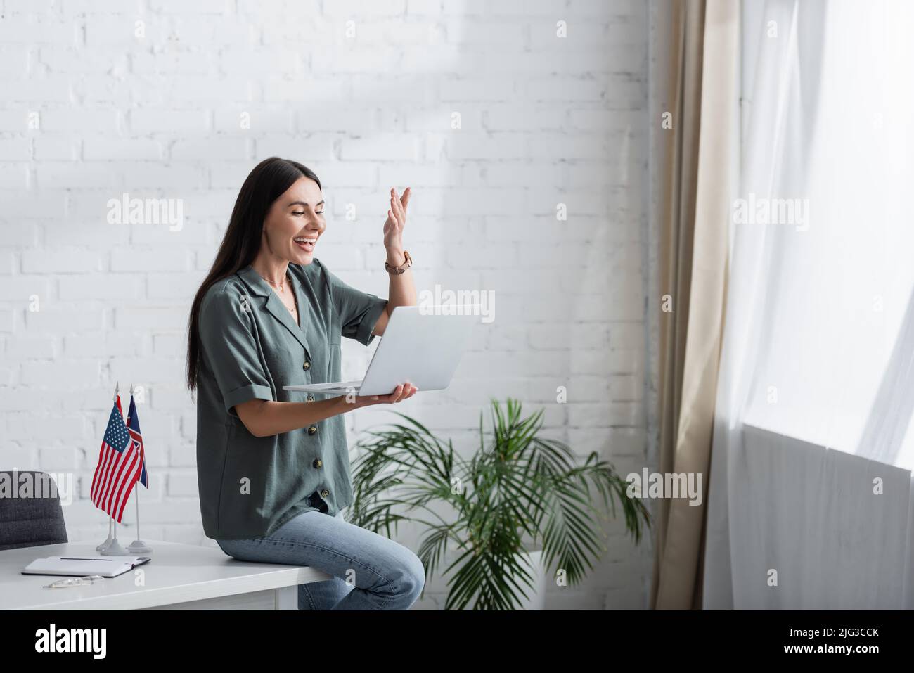 Cheerful teacher holding laptop during online lesson near flags on ...