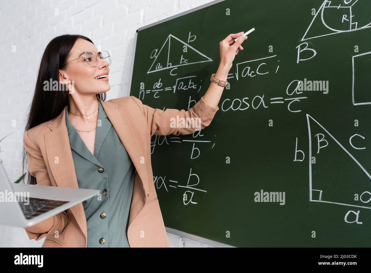 Teacher holding laptop during online lesson near chalkboard with math ...