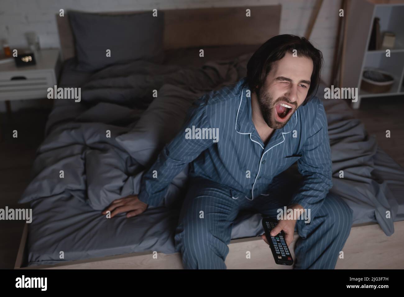 Tired man yawning and holding remote controller in bedroom at night ...
