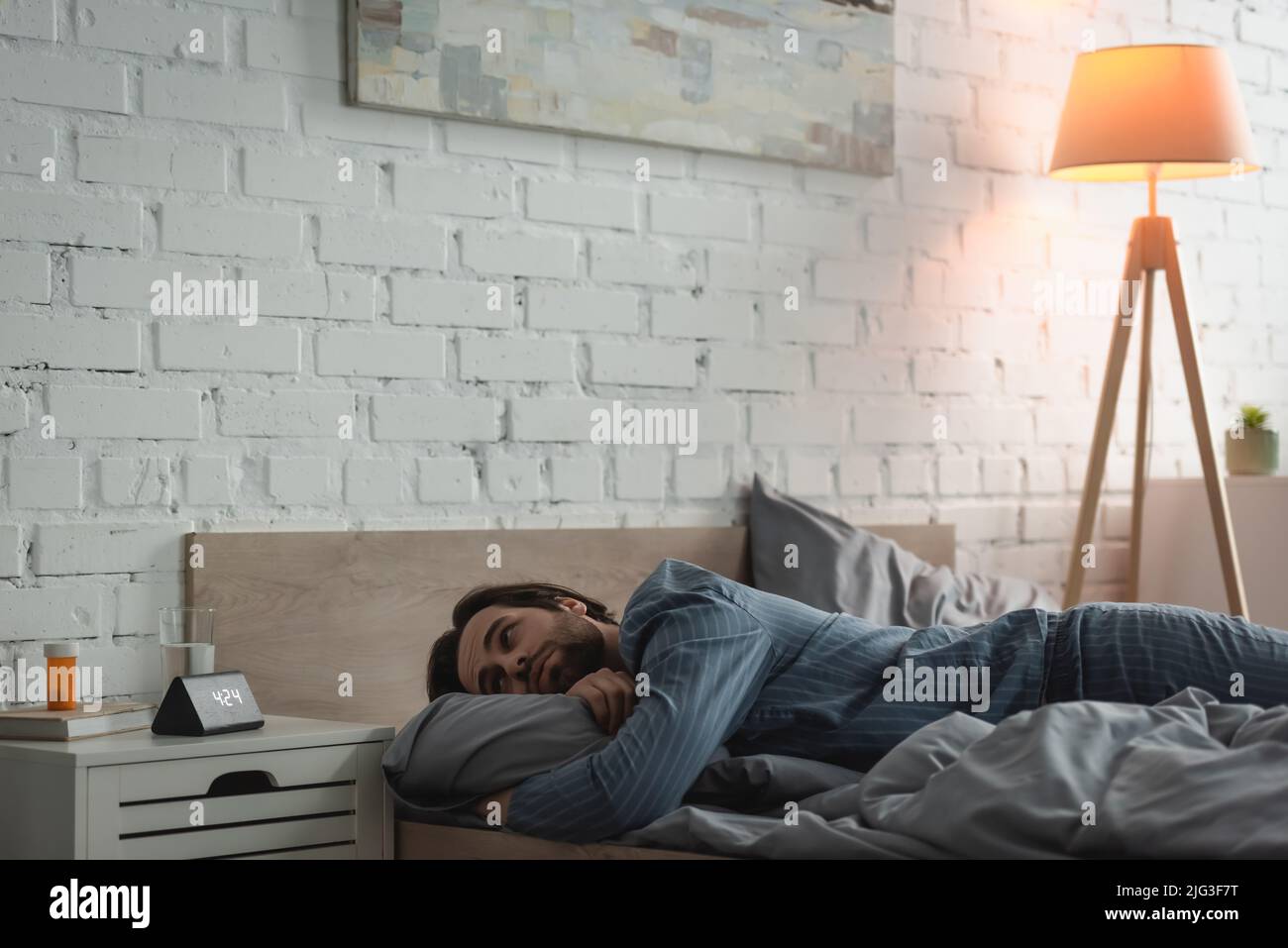 Young man looking at clock while suffering from insomnia on bed in ...