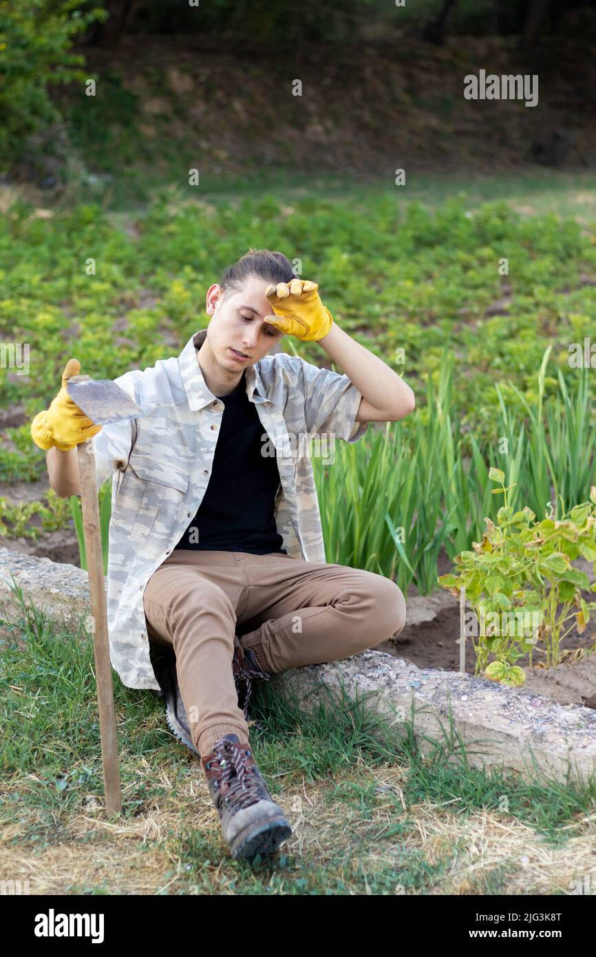 Vertical view of young gardener tired sitting with a hoe after working on the garden cultivating ...