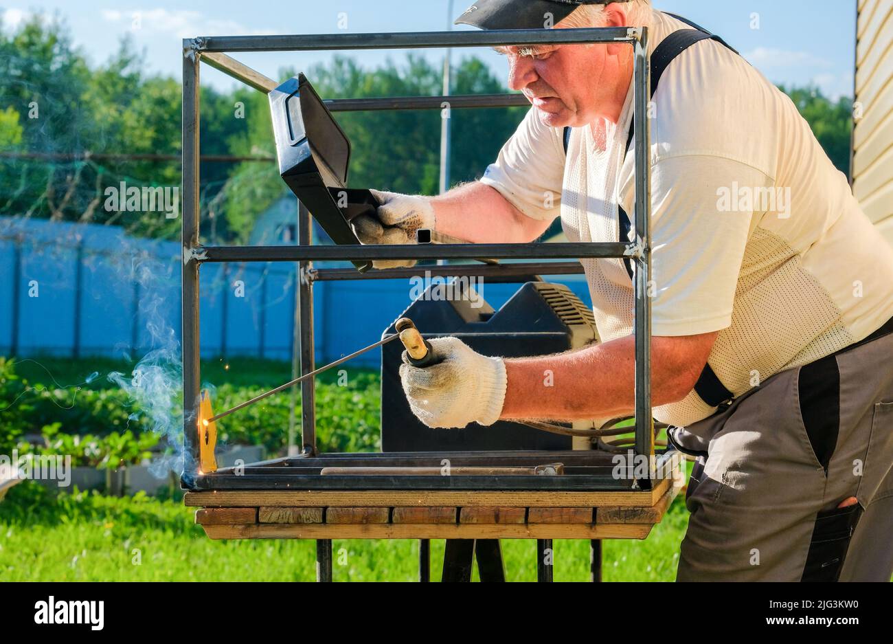 A senior gray-haired man welds a metal structure with electric arc ...