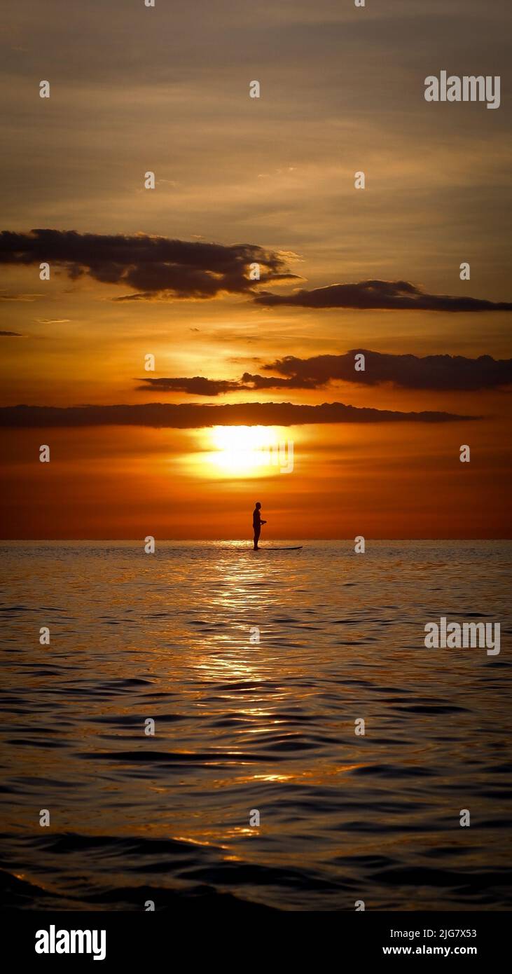 A silhouette of a man paddle boarding in the sea during a scenic sunset ...