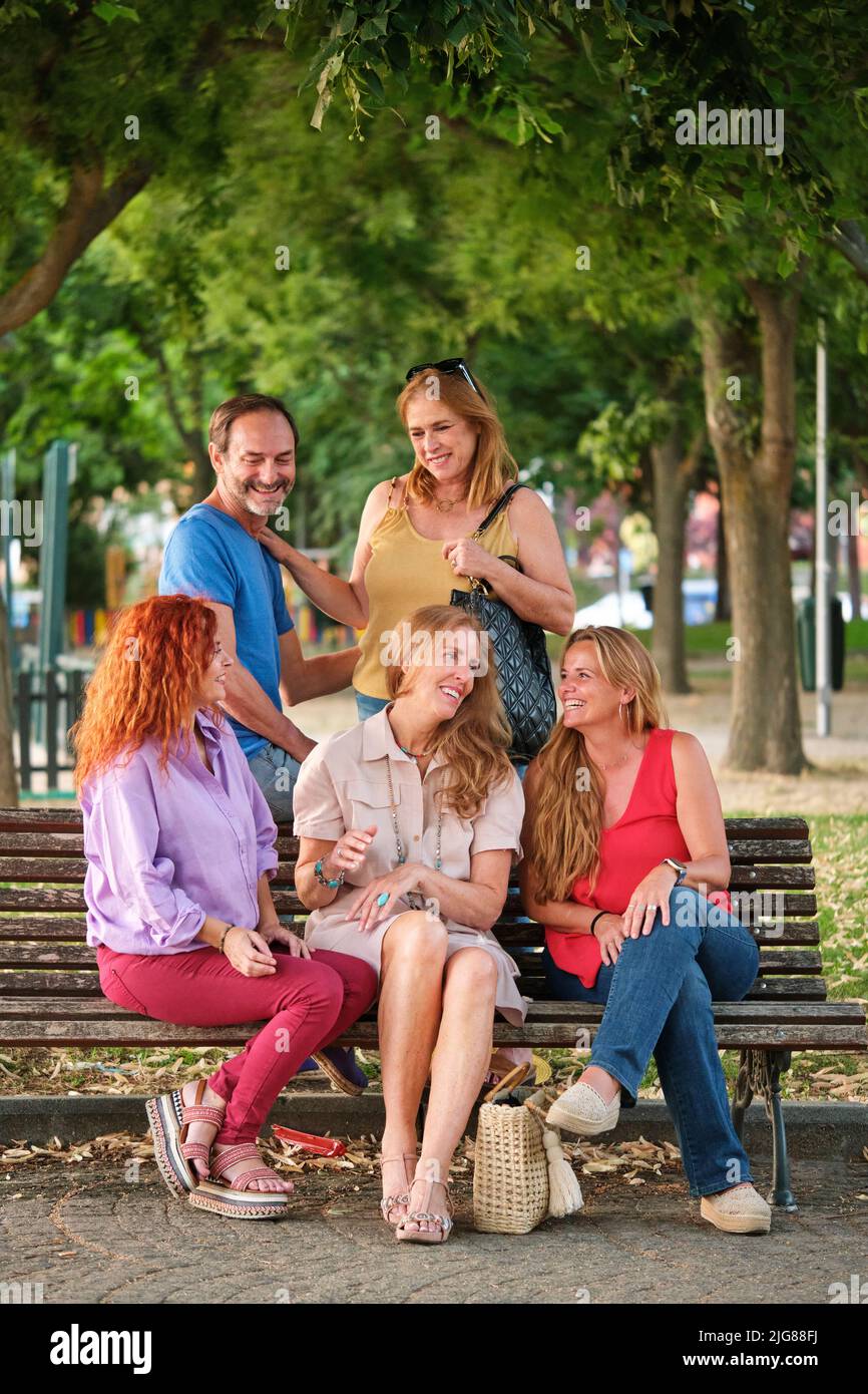 Five mature adults talking and laughing sitting on a bench in a park ...