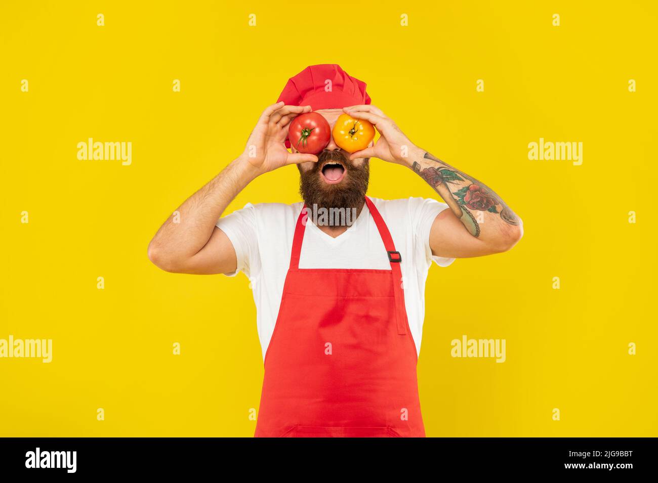 Happy guy in cooking apron and toque holding tomatoes on eyes yellow ...