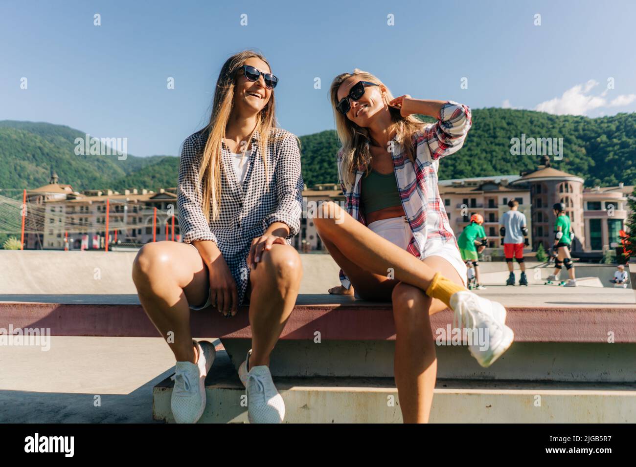 Two young carefree tanned women are sitting on a bench in a skatepark ...