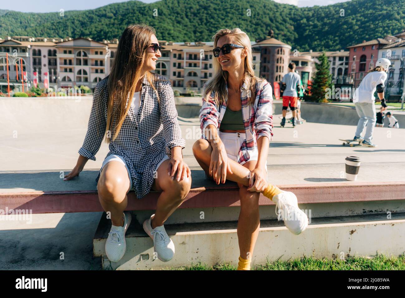 Two young Caucasian tanned women are sitting on a bench in a skatepark ...