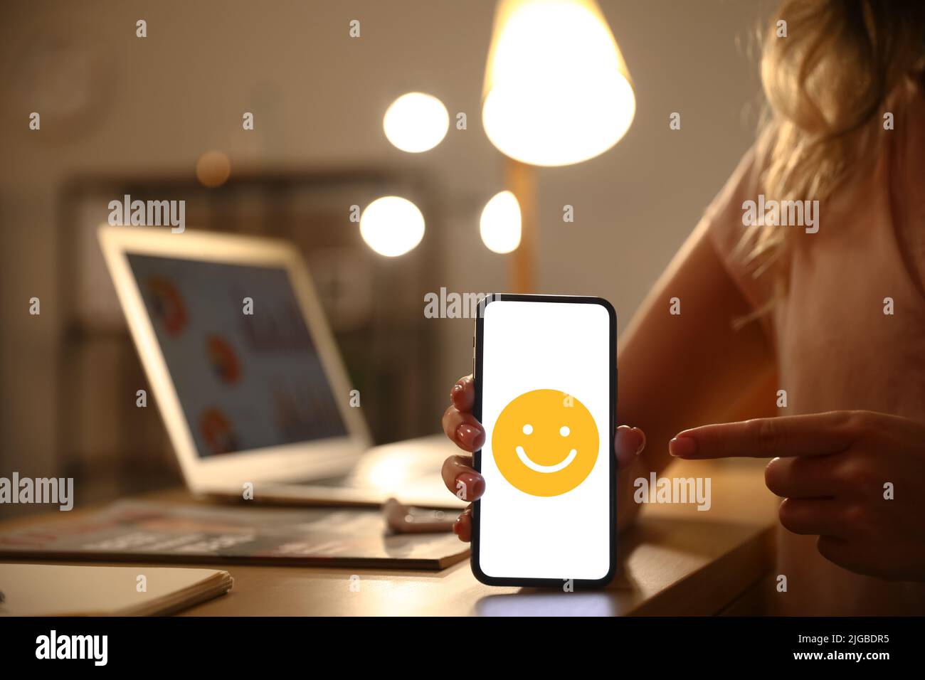 Woman holding phone with happy emoticon on screen at home in evening ...