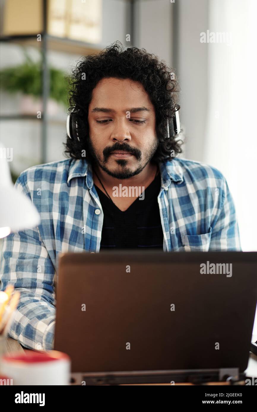 Portrait of Indian software developer with curly hair listening to music in headphones when ...