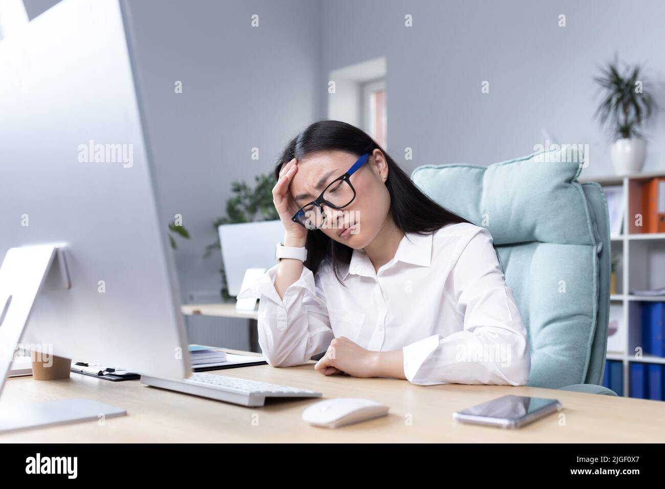 Exhaustion at work. A young Asian woman in glasses holds her head ...