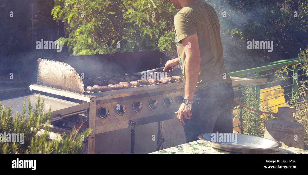 Young guy cooking sausages over open fire while camping in nature. BBQ ...